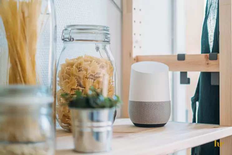 In Google Home smart speaker sit op in houten planke neist in pot fol mei noedels yn in keuken.