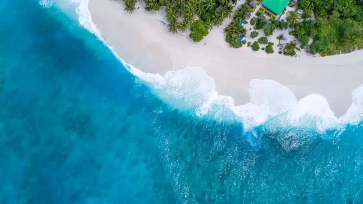 Globoko modri valovi prihajajo na obalo blizu hiše, ki leži sredi zelenih dreves na Maldivih.